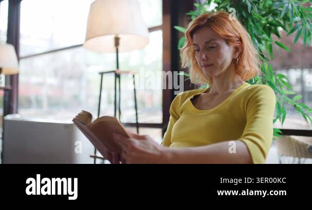 In a cafe, a woman in yellow reads a book near a window and an indoor ...