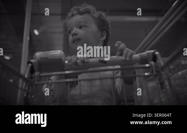 Black-and-white view of a toddler boy standing in a metal shopping cart ...