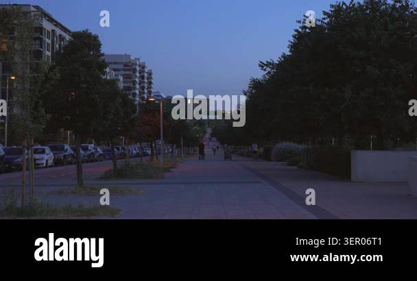 People walk along a tree-lined urban promenade at dusk. Residential ...