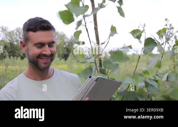 A man smiles while holding a tablet and looking at a sapling in a field ...