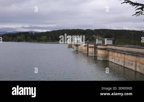A wide shot of a concrete dam with water on one side and a forest on ...