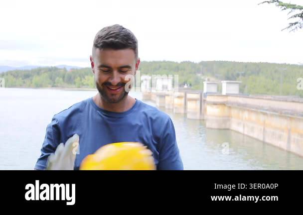 A man wearing gloves puts on a yellow hard hat in front of a dam and a ...