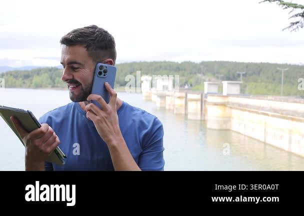 A man is talking on his phone and holding a tablet in front of a dam ...