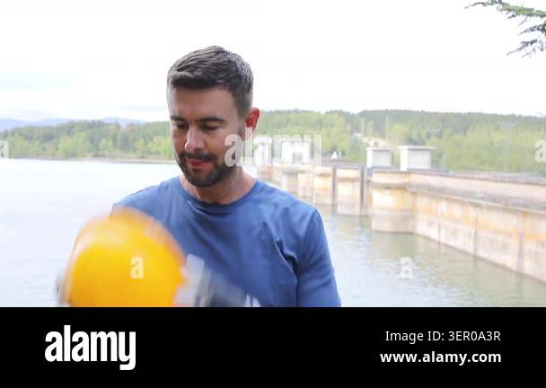 A man wearing gloves puts on a yellow hard hat in front of a dam and a ...