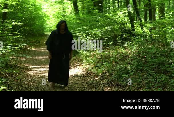 Monk in a dark cloak walks barefoot along a forest road among green ...
