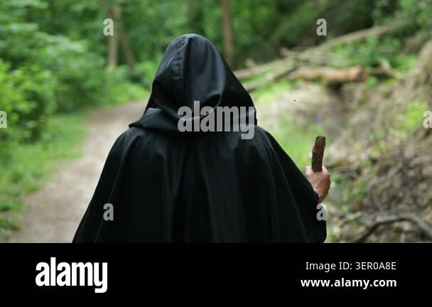 Monk in a dark cloak walks along a natural trail using a wooden staff ...