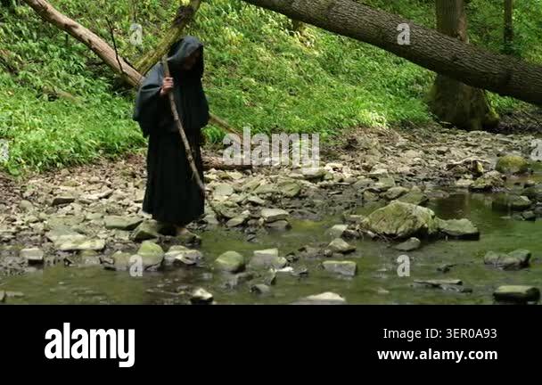 Monk in a dark cloak carefully crosses a rocky forest stream using a ...