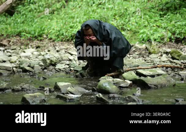Monk in a dark cloak quenches his thirst with clean water from a forest ...