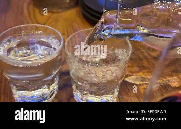 Clear water is poured from a glass pitcher into two drinking glasses on ...