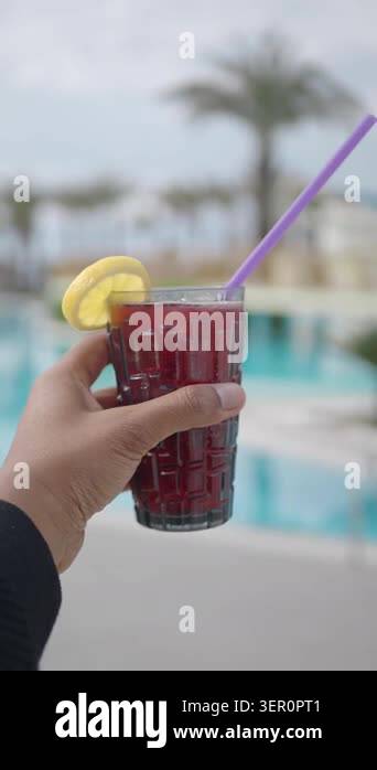A person holds a drink with a lemon slice near a pool. The sun shines ...