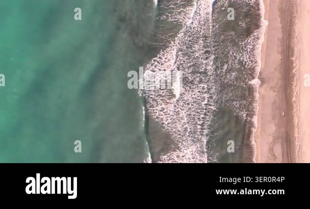 Drone looks straight down as waves break and foam washes over a wide ...