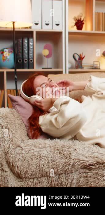 Young woman on home sofa listening to music in wireless headphones with ...