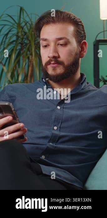Lebanese man sitting on sofa uses mobile phone smiles at home living ...