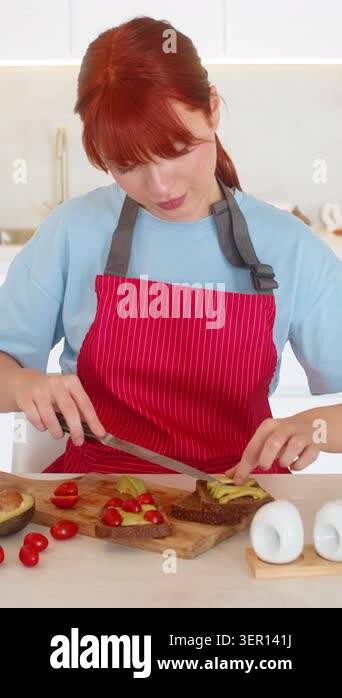 Young woman at home in kitchen wearing apron prepares avocado toast ...