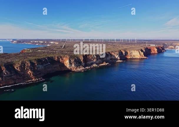 Cliffs rise from the blue water while wind turbines stand in a line on ...