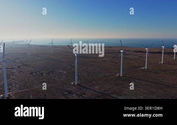 Wind turbines are arranged in a line on flat land next to the ocean ...