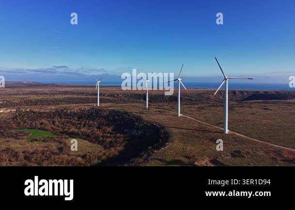 Wind turbines are positioned on a hillside overlooking the ocean. The ...