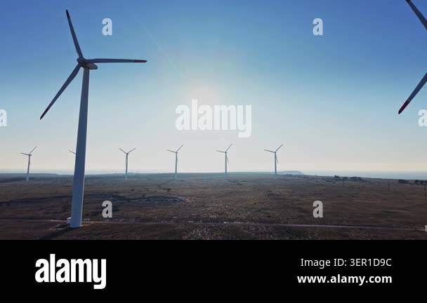 Wind turbines turn slowly in an open field under a clear blue sky. The ...