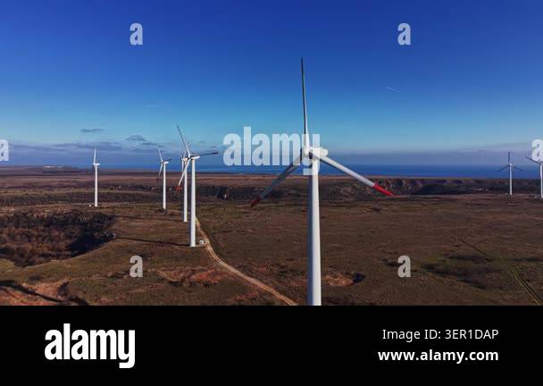 Wind turbines rotate in a field by the sea. The landscape features open ...