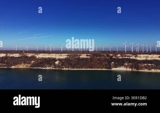 Wind turbines are lined up near a cliff overlooking a body of water ...
