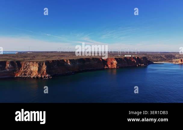 A rocky coastline meets blue water. In the background, wind turbines ...