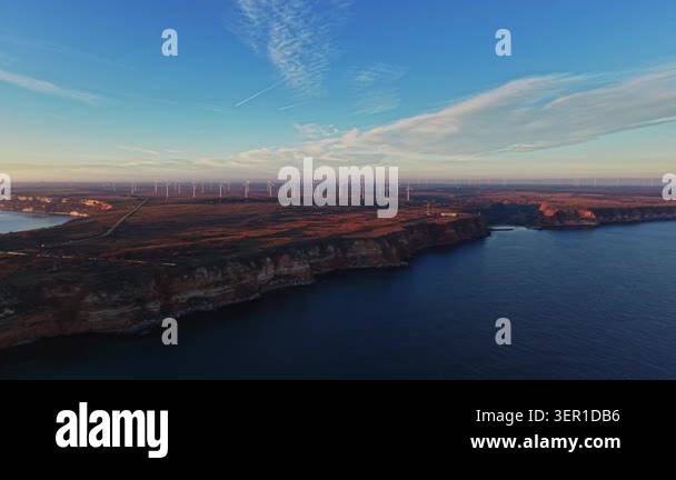 Wind turbines stand on a cliff near the water. The sun sets on the ...