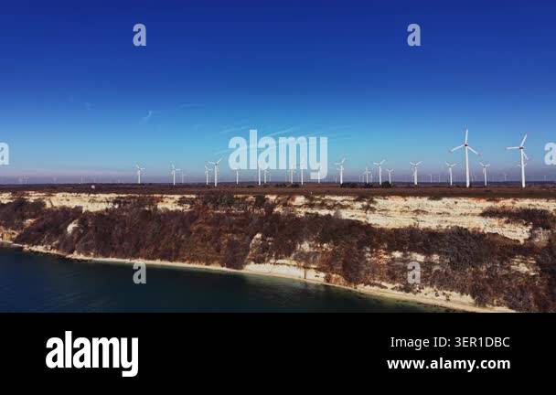 Wind turbines stand on the edge of a cliff overlooking the water. The ...
