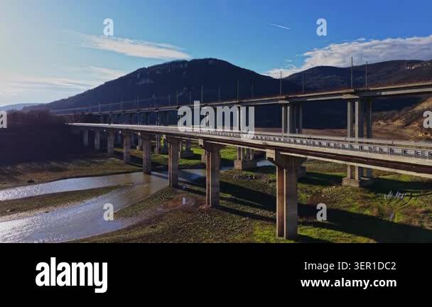 A large bridge stands over a river with hills in the background. The ...