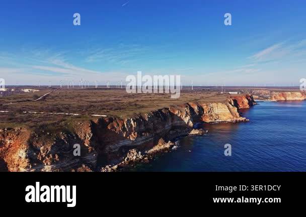 Cliffs meet the sea under a clear sky. Wind turbines stand on the land ...