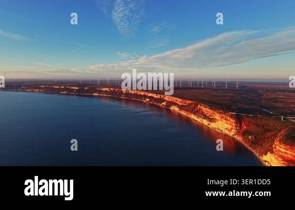 Wind turbines stand on the cliffs overlooking the water. The sun is ...