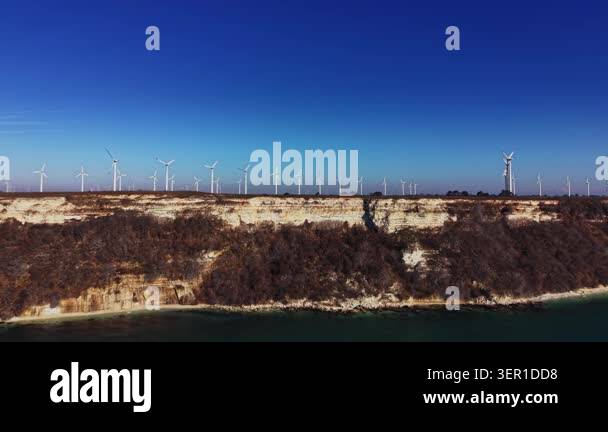 Wind turbines are lined along a cliff edge by the coast. The sky is ...