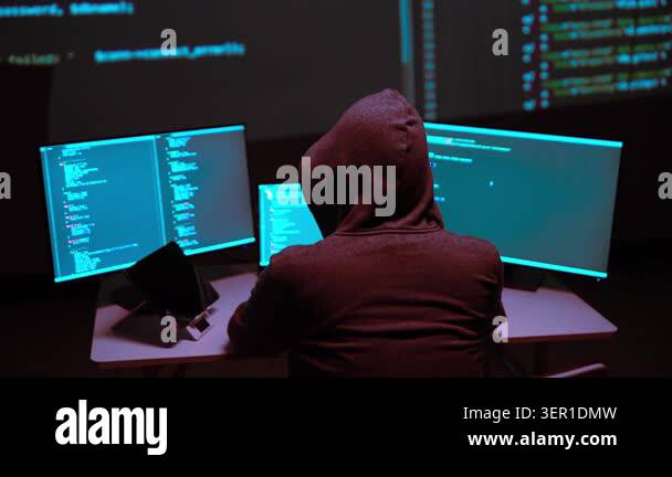 A solitary figure dressed in a hoodie sits in front of multiple screens ...
