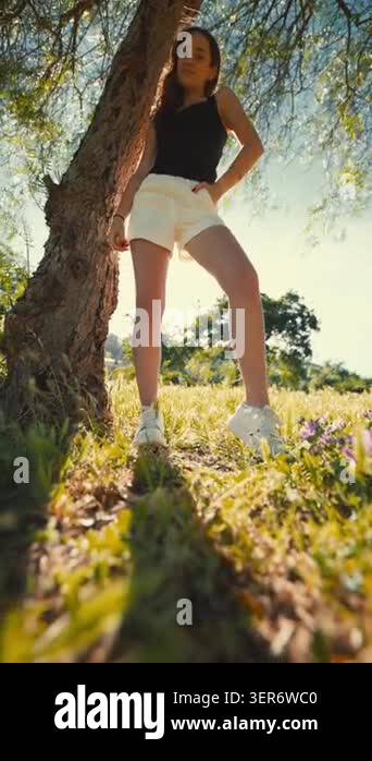 A woman stands confidently beside a tree in a sunny field filled with ...
