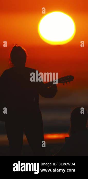 A person plays guitar on the beach during sunset. People watch as the ...