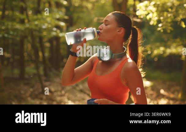 Young woman drinking water after jogging in forest at sunset and ...
