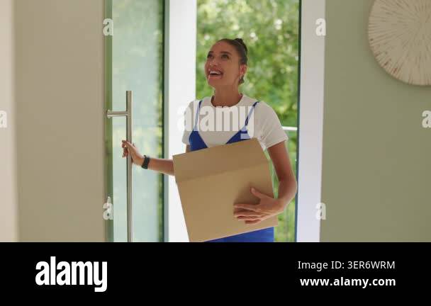 Smiling young woman opening front door while carrying moving box ...