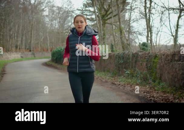 Cardio focused female running on curved rural road surrounded by forest ...