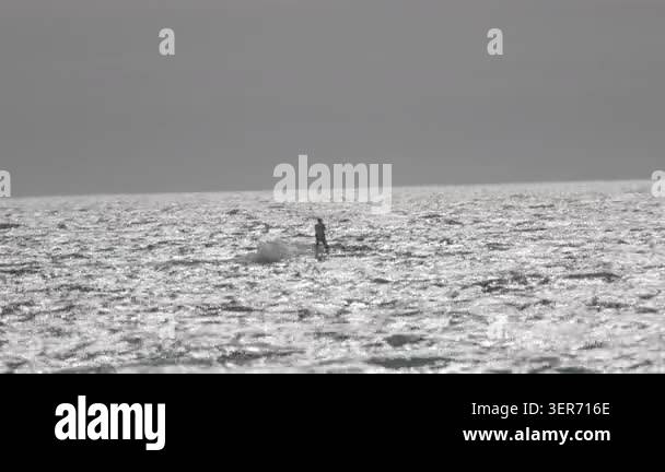 A person stands on a paddleboard in the ocean under bright sunlight ...