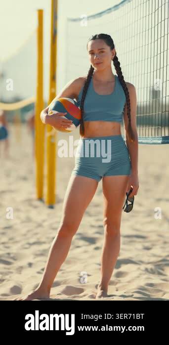 A young woman stands on a beach volleyball court with a ball and ...