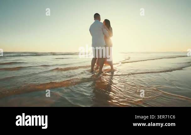 A couple stands close together in shallow water at the beach. The sun ...
