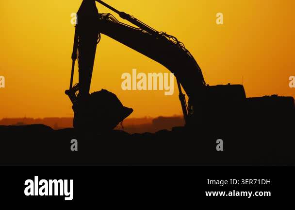Excavator operates at sunset, lifting and moving earth in a ...