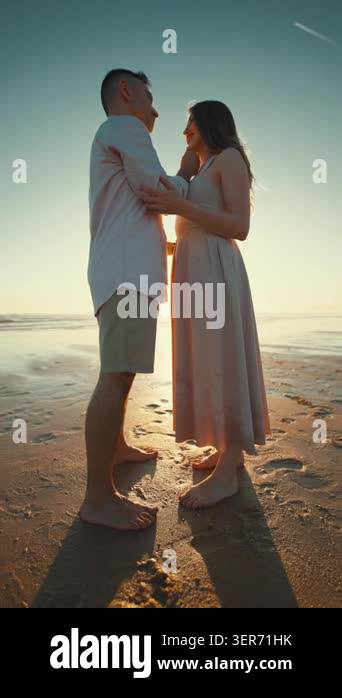 A couple stands barefoot on a sandy beach as the sun sets behind them ...