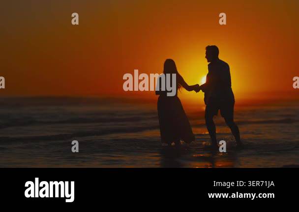 Two people walk on the beach at sunset. They hold hands and smile as ...