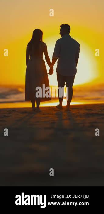 A couple stands on the beach holding hands as the sun sets behind them ...