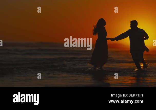 A couple runs along the shore at sunset, splashing water as they play ...