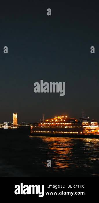 A ferry moves through water at night. City lights shine brightly in the ...