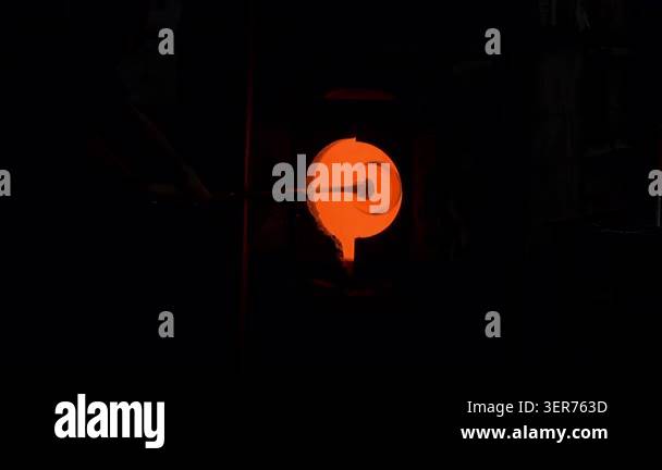 A craftsman shapes molten glass on a rotating rod in a hot, dark ...