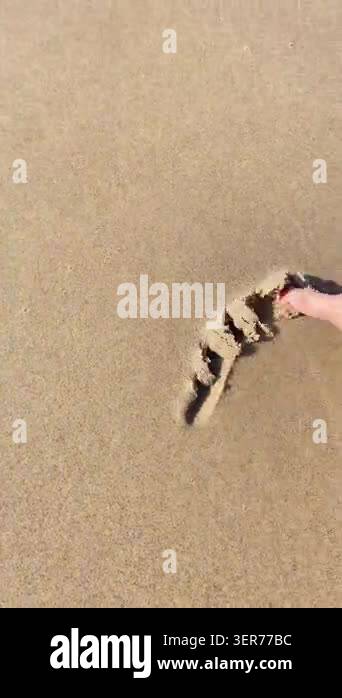 Vertical video of a hand drawing a heart shape in the sand near the ...