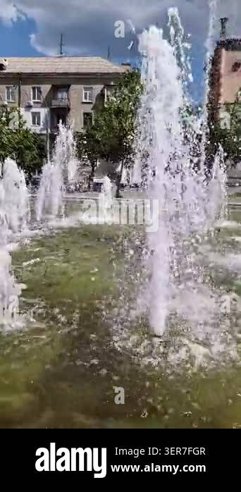 Bright water jets erupt from a stone fountain in a sunny city square ...