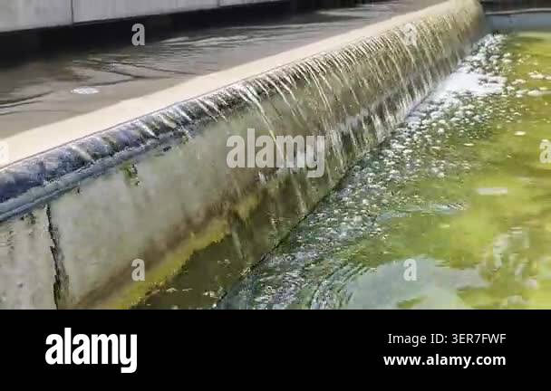 Water flows steadily over a smooth concrete ledge into a rippling pool ...